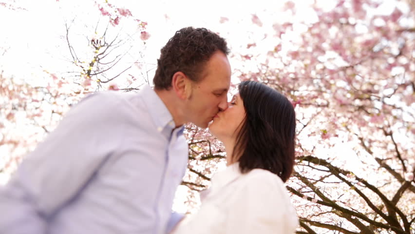 Mature Couple Standing Under Cherry Tree First Looking At Camera Than At Each Other For A Kiss